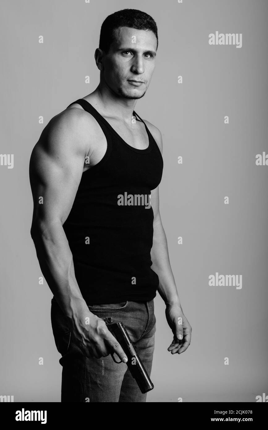 Studio shot of young muscular Persian man standing while holding gun ...