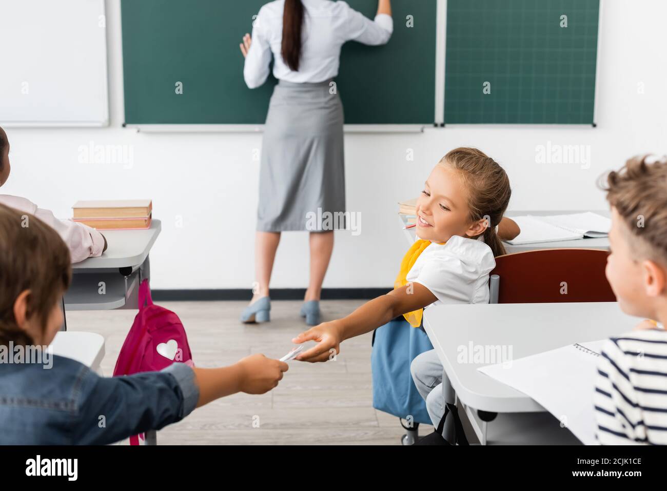 selective focus of pupil passing note to schoolgirl while teacher ...