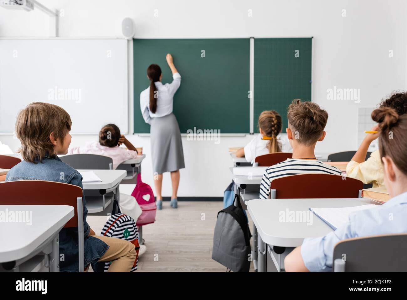 back view of teacher writing on chalkboard near multicultural pupils in ...