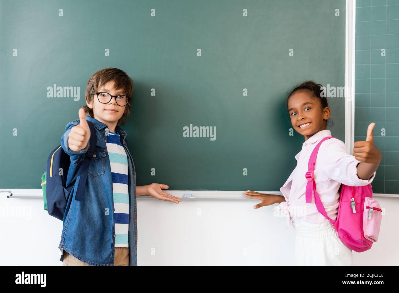 Multiethnic schoolchildren showing thumbs up at camera near chalkboard ...