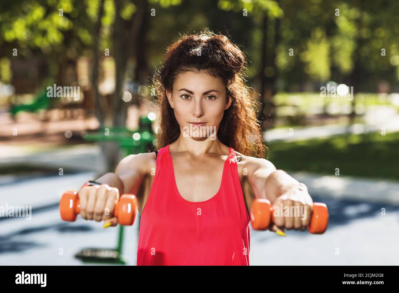 Young woman exercises shoulders with dumbbells outdoors on a warm sunny ...