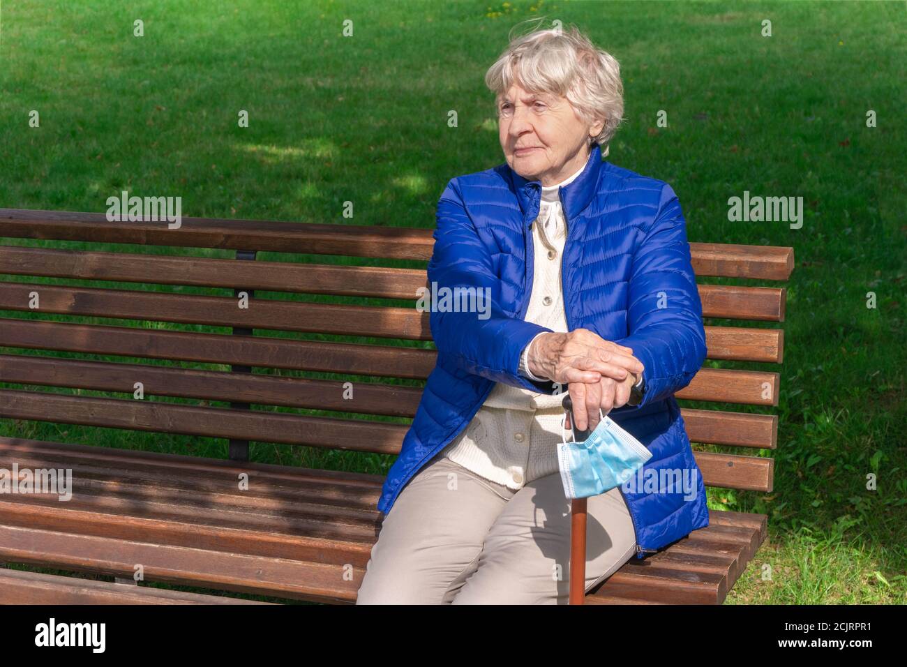 Old woman with protective face mask on cane sits on bench in park ...