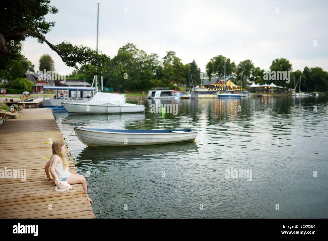 Cute young girl sitting on a wooden platform by the lake dipping her ...