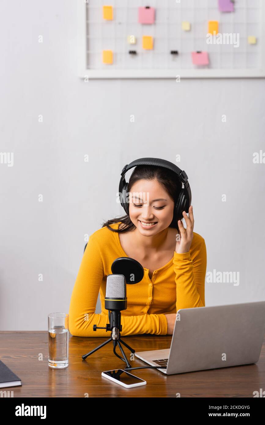 young asian radio host touching wireless headphones while sitting near ...