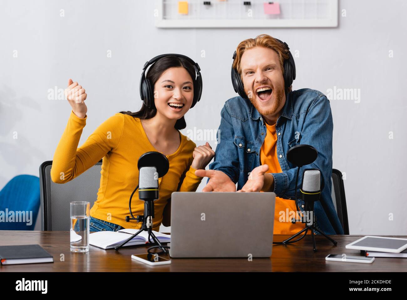 excited broadcaster pointing with hands at laptop near asian colleague ...