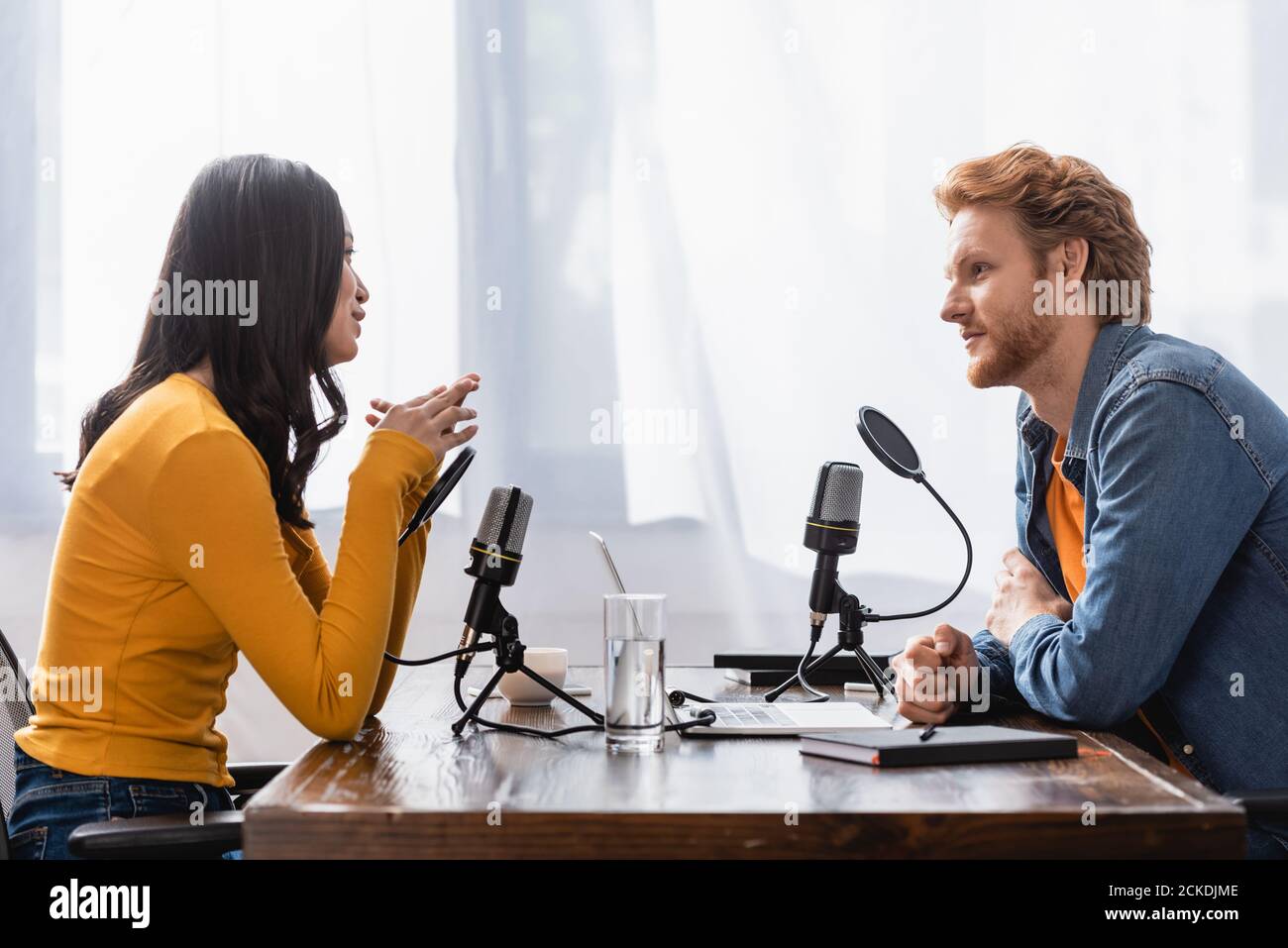 side view of young asian woman talking to redhead broadcaster during ...
