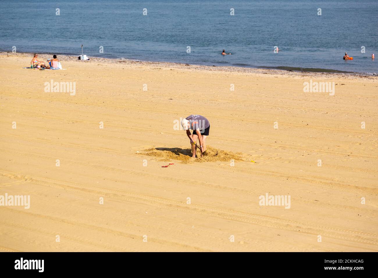 A middle aged man digging in the sand on a beach with a bucket and ...