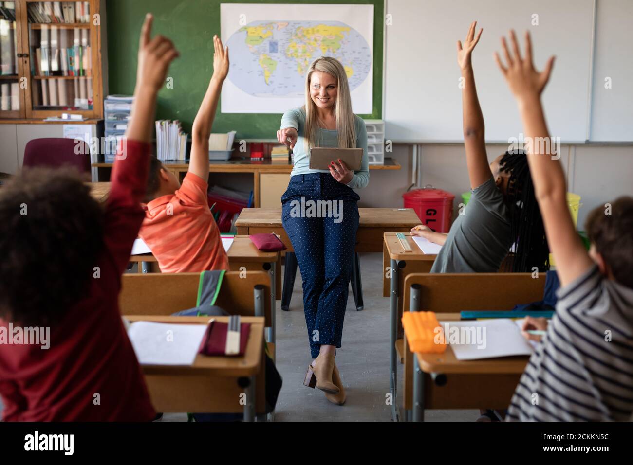 Female teacher pointing towards kids raising their hands in class at ...