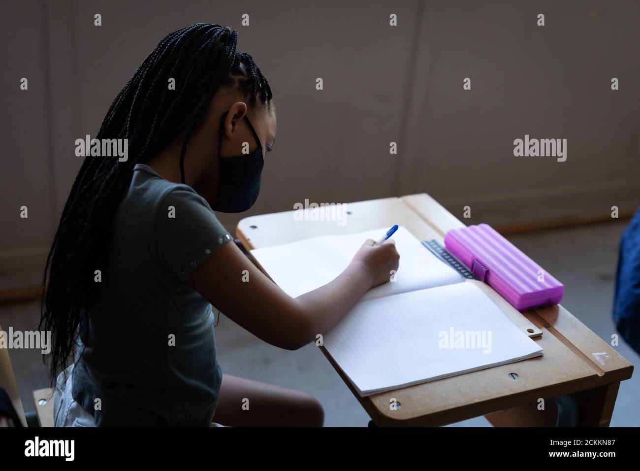 Girl wearing face mask writing while sitting on her desk at school ...