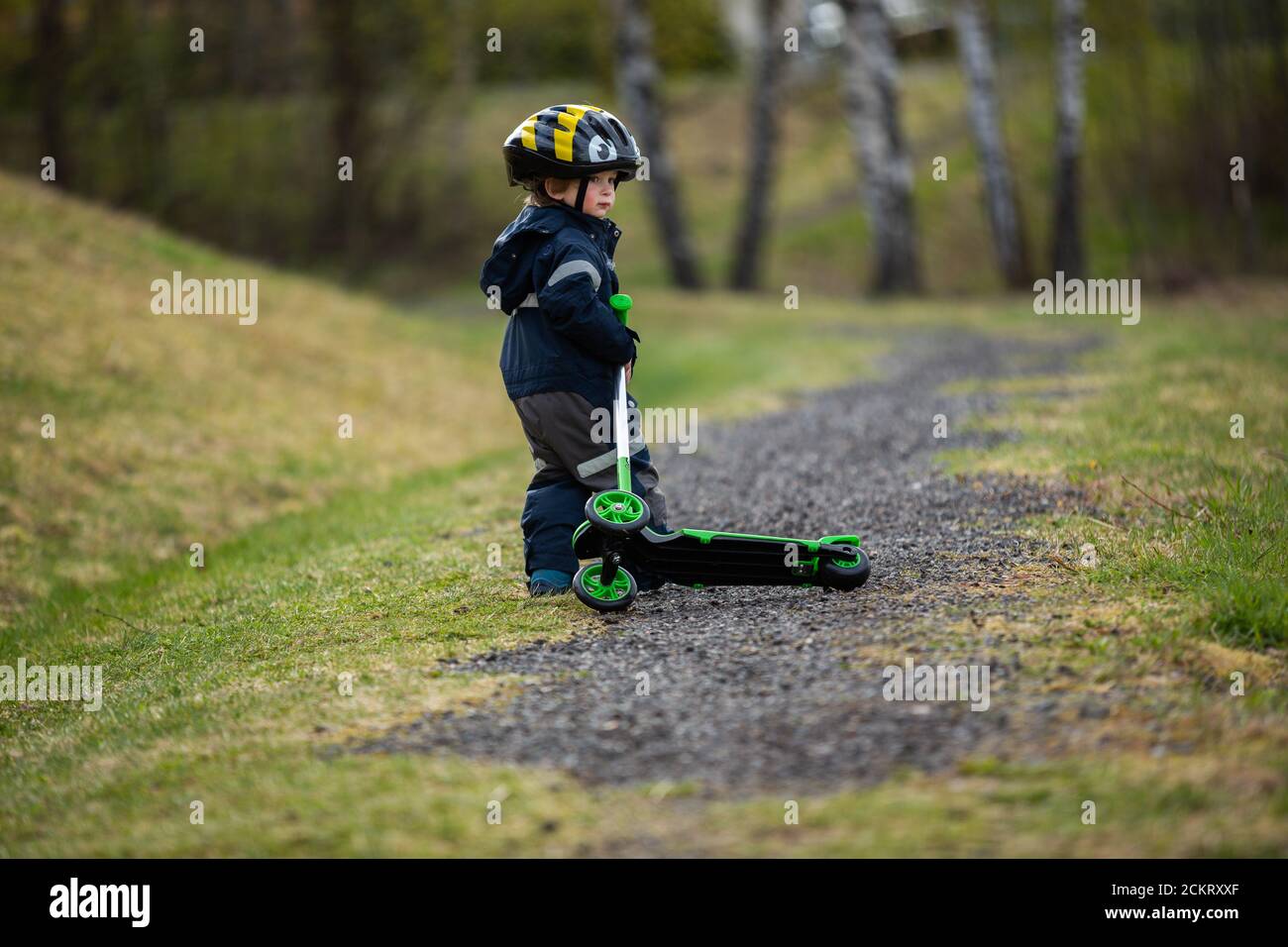 Young boy wearing a bike helmet dragging a kick scooter up a pebbled ...