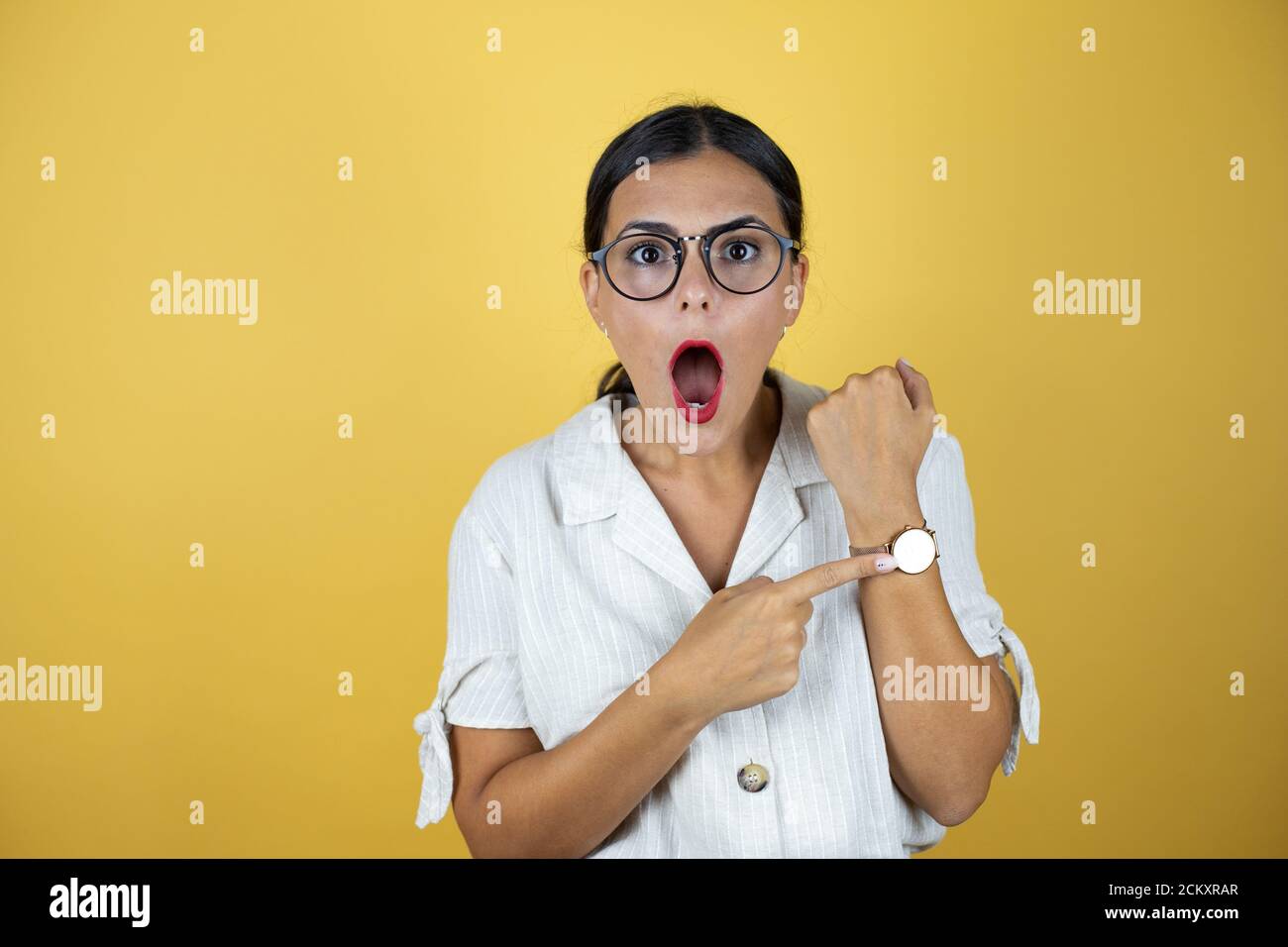 Beautiful woman over yellow background surprised and pointing the watch ...