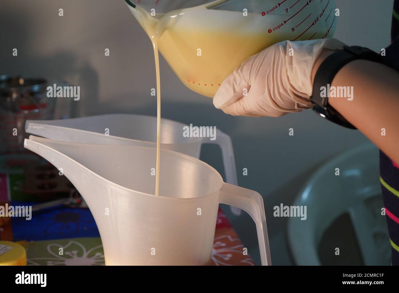 Gloved hand with watch pouring white liquid from glass jar into a white ...