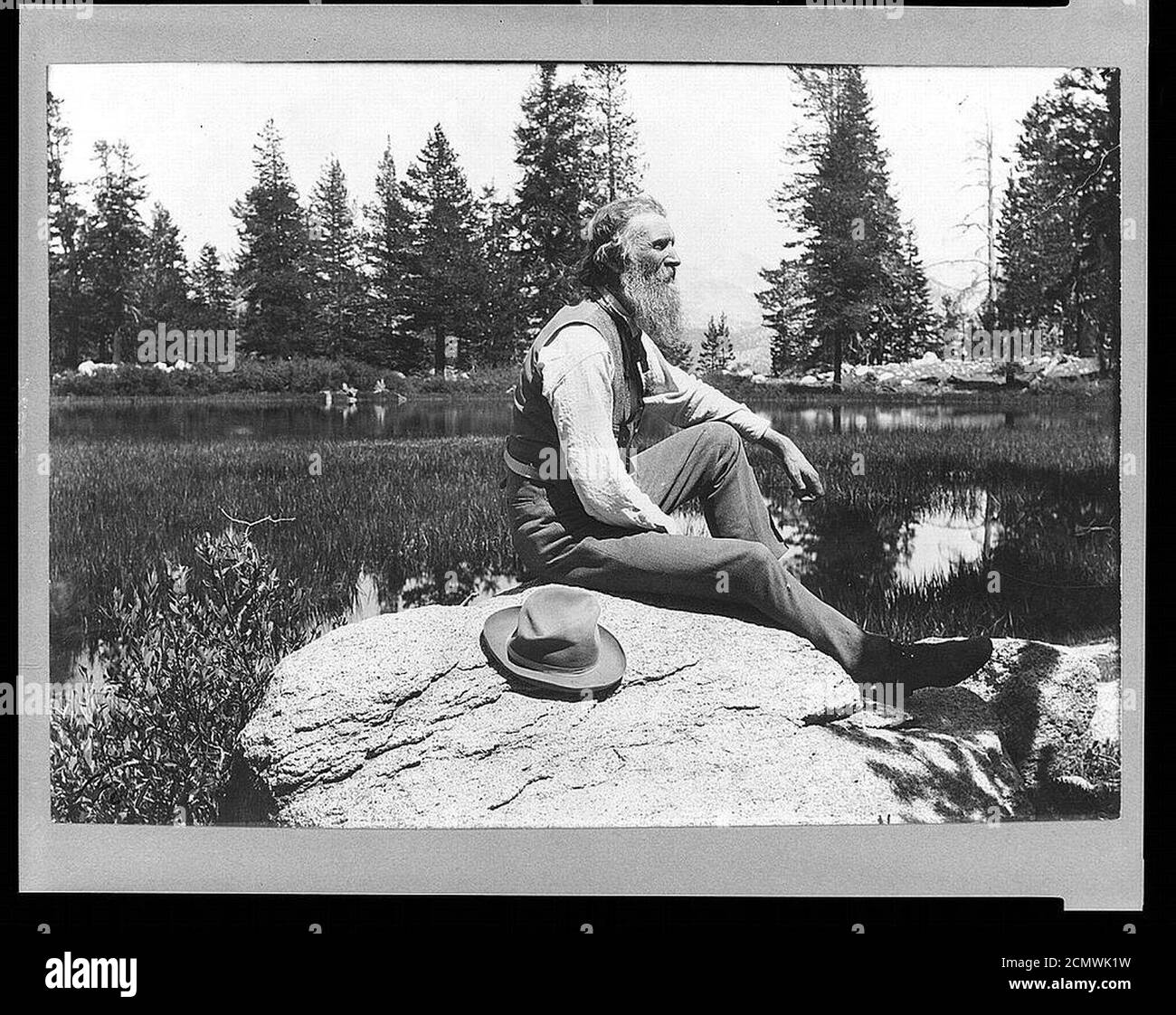 John Muir, full-length portrait, facing right, seated on rock with lake ...