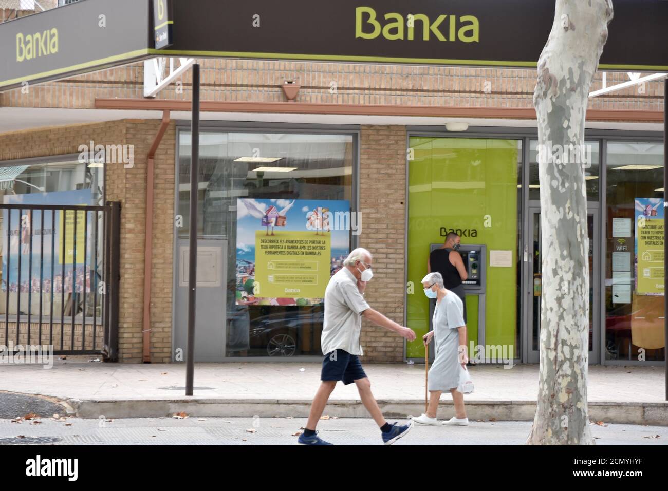 People wearing facemasks walk past the Bankia logo seen on the facade ...