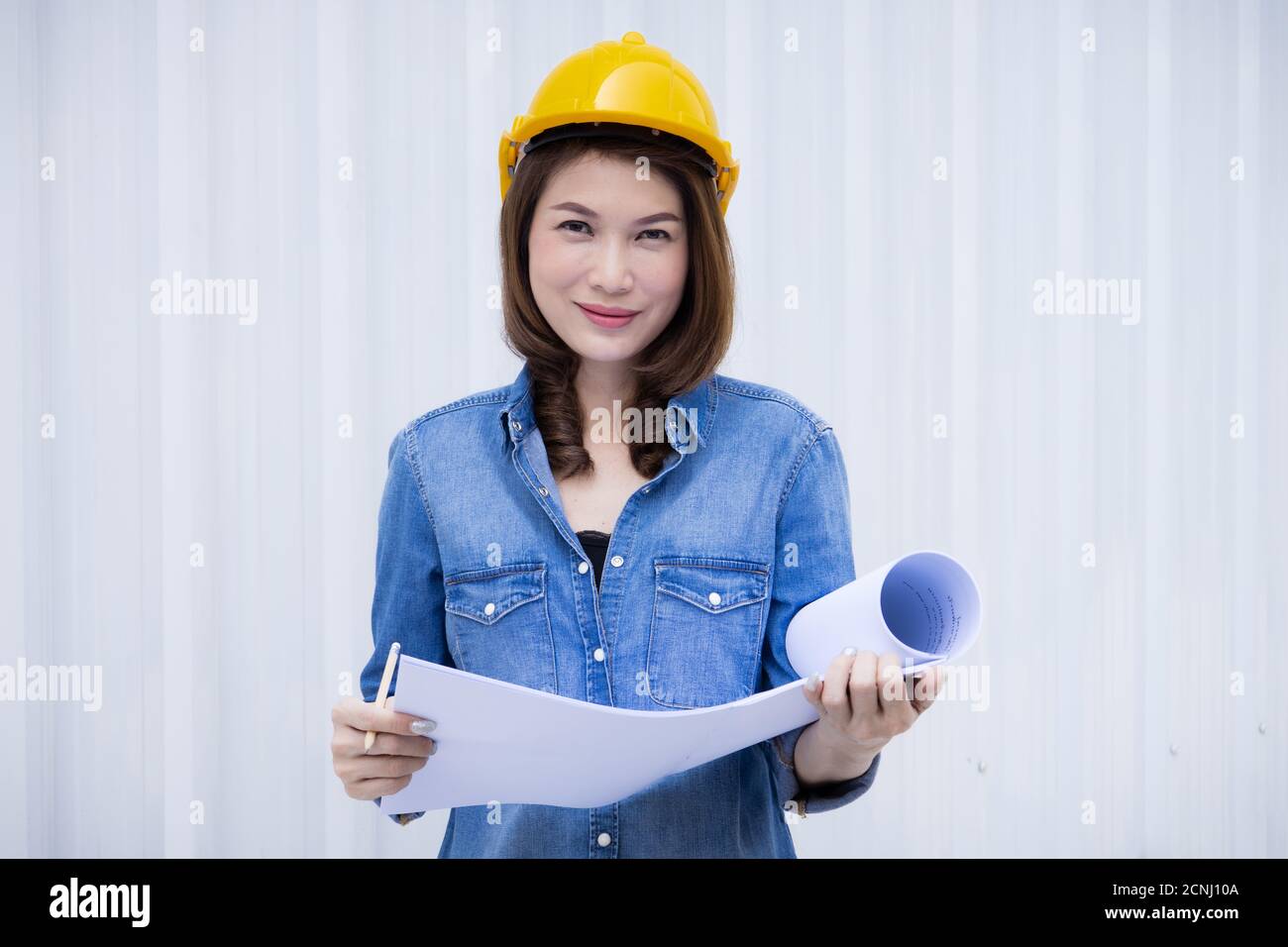 Beautiful Asian female engineer in jeans dress wearing yellow safety hard hat doing job at ...