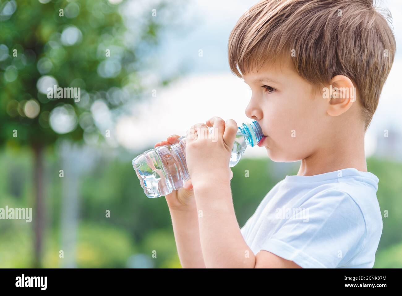 Handsome boy drinks clean water from a bottle on the street in summer ...