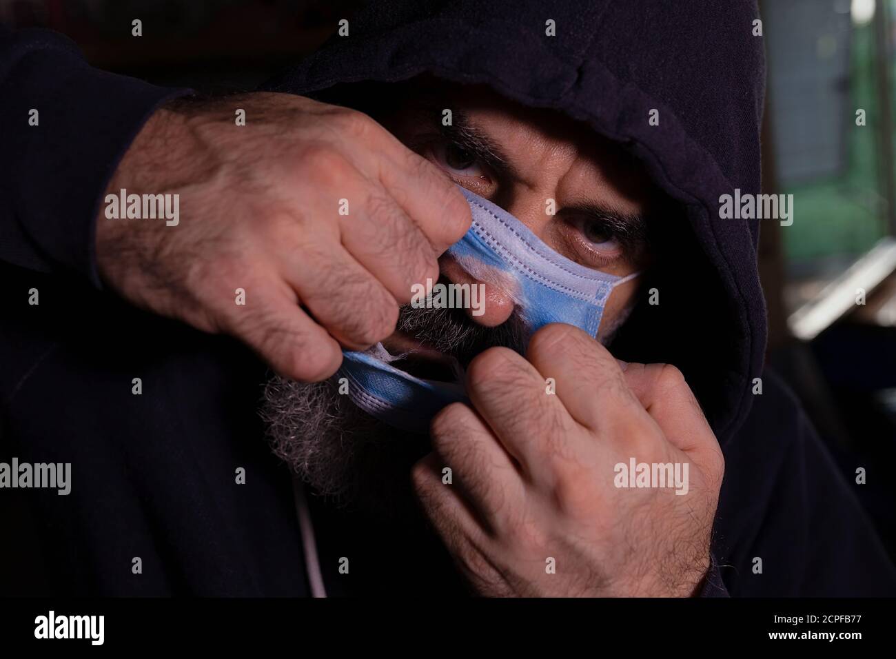 Bearded man in dark hoodie ripping blue surgical mask on his face Stock ...