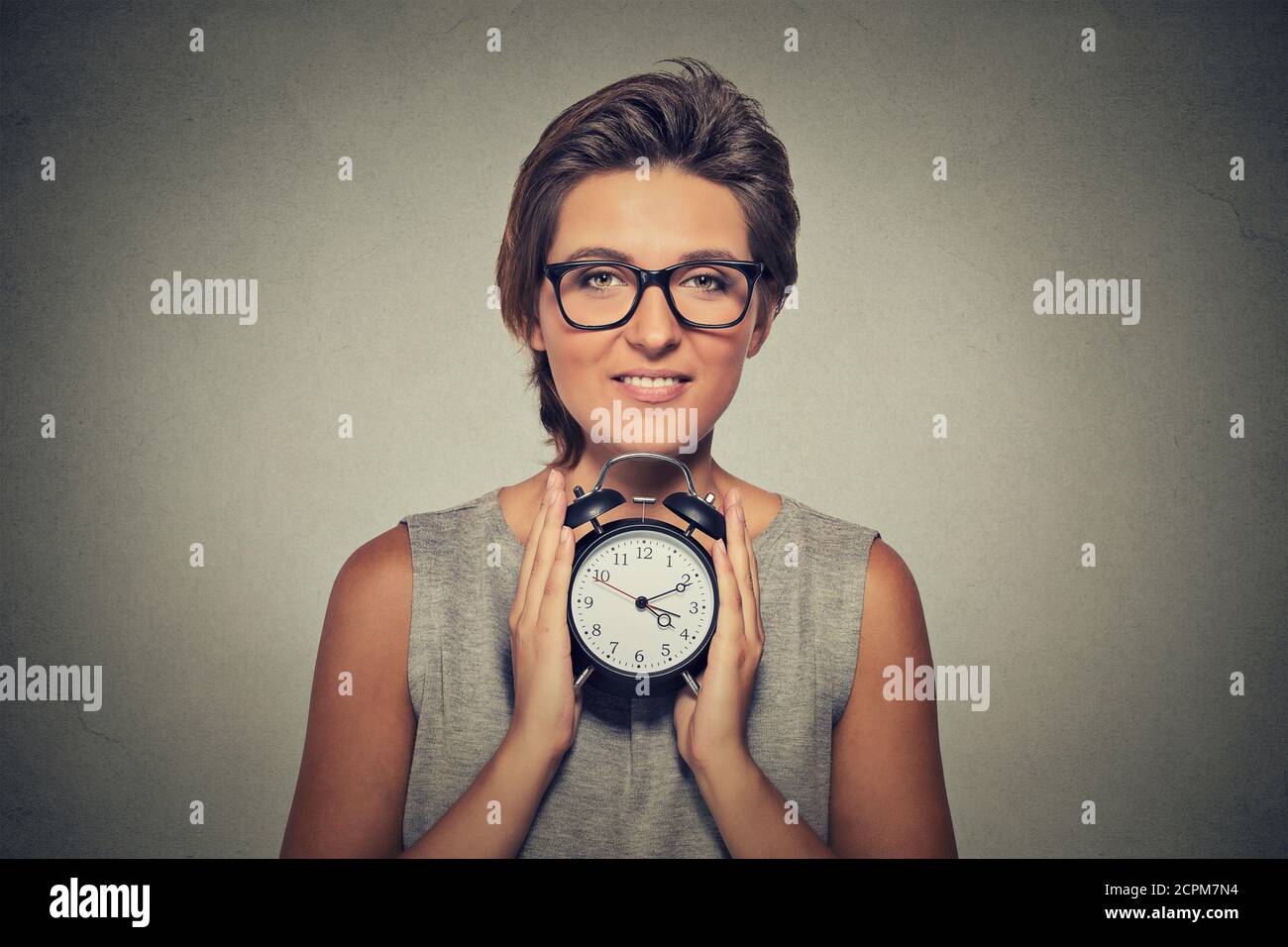 young smiling woman with alarm clock isolated grey wall background ...