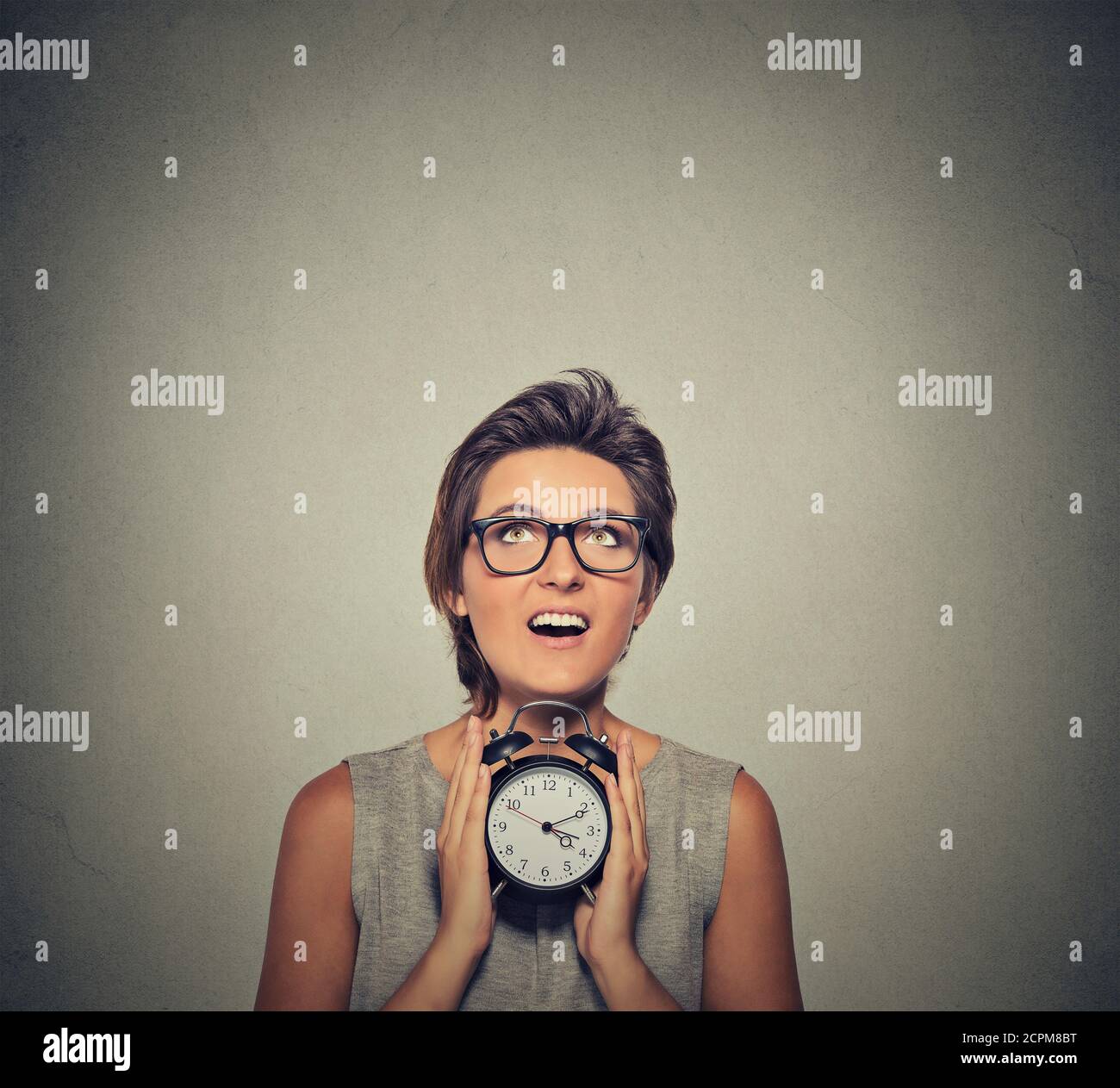 young smiling woman with alarm clock looking up isolated on grey wall ...