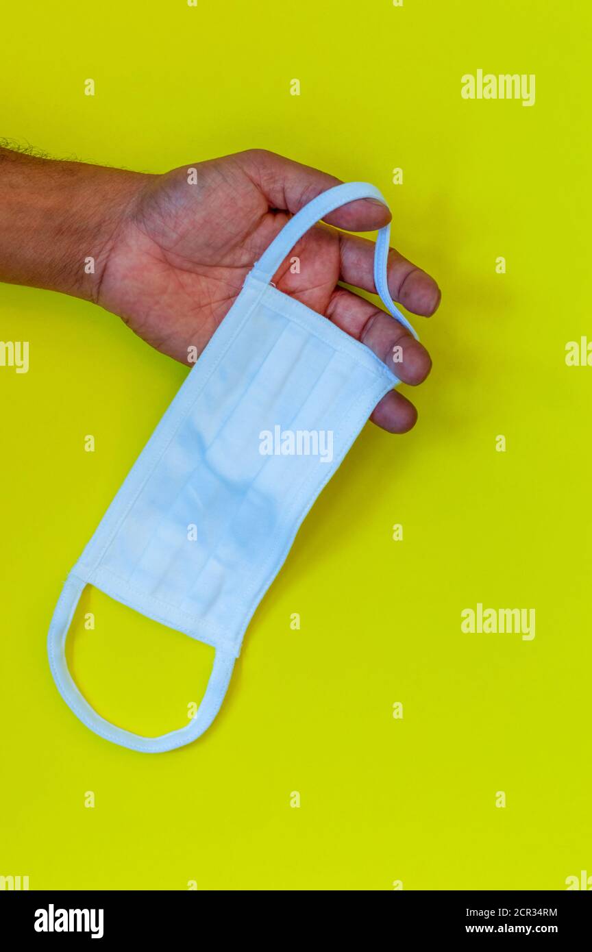 Left hand holding a white face mask on a yellow background. Public ...