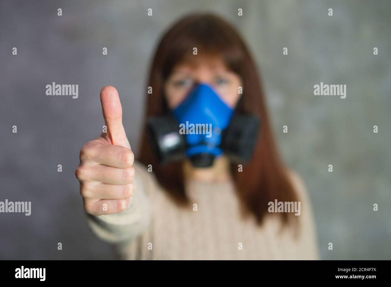 Young woman in respirator with blurred face shows thumb up on grey ...