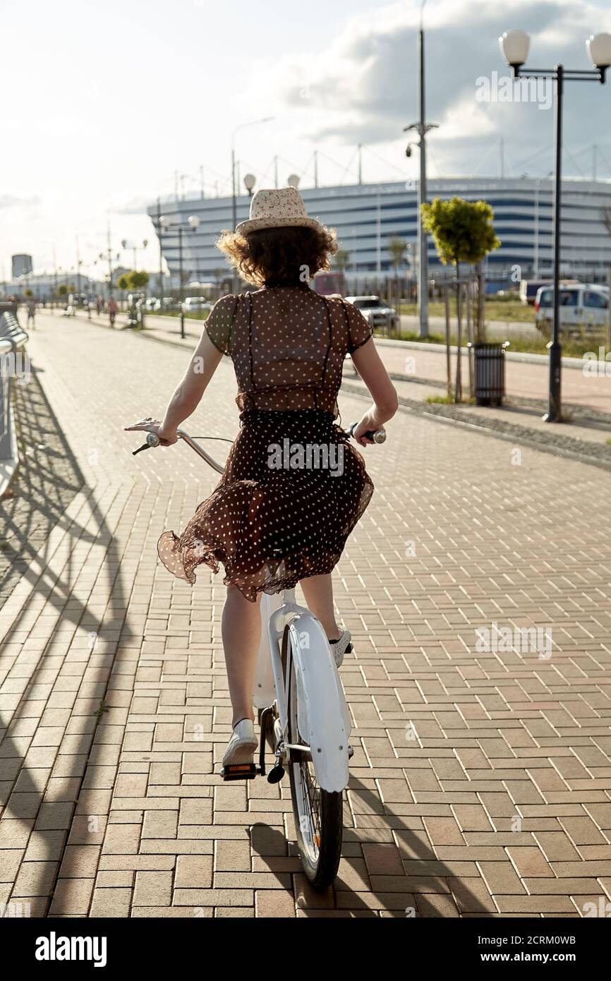 young curly hair woman in hat and dress moves on bike near river at ...