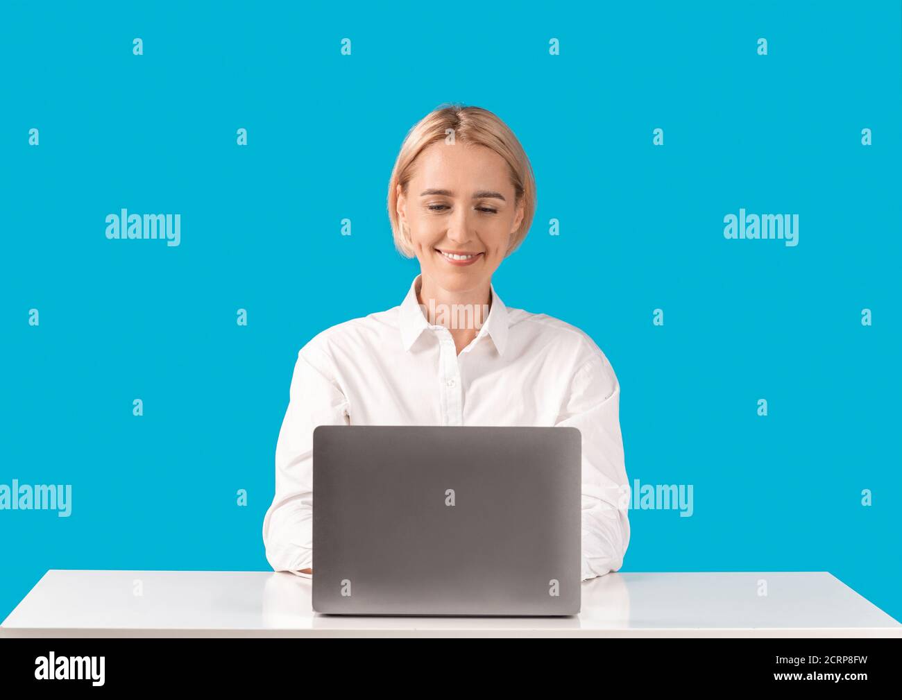 Positive business lady using laptop computer at her desk on blue studio ...