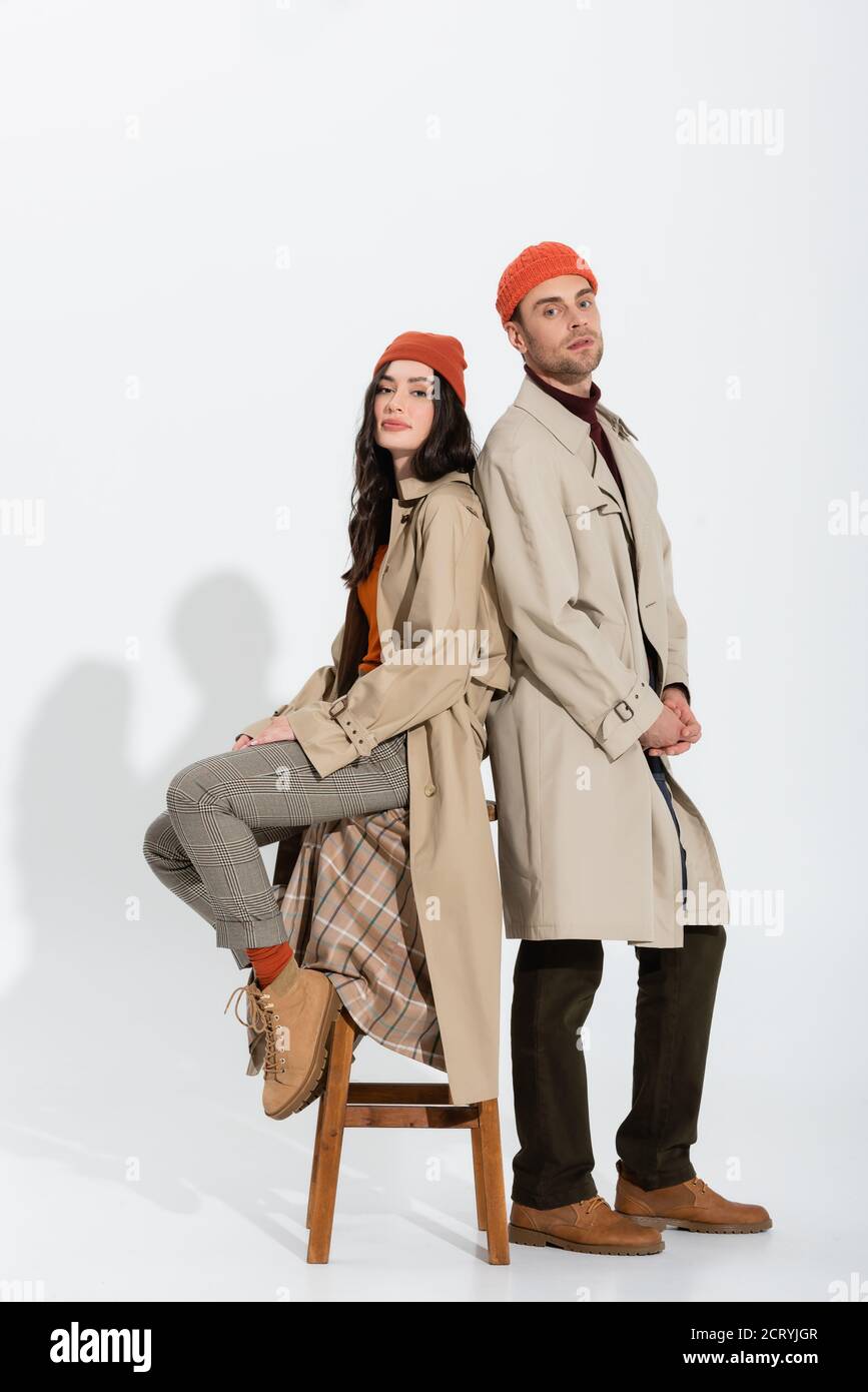 full length of stylish woman in trench coat sitting on stool and ...