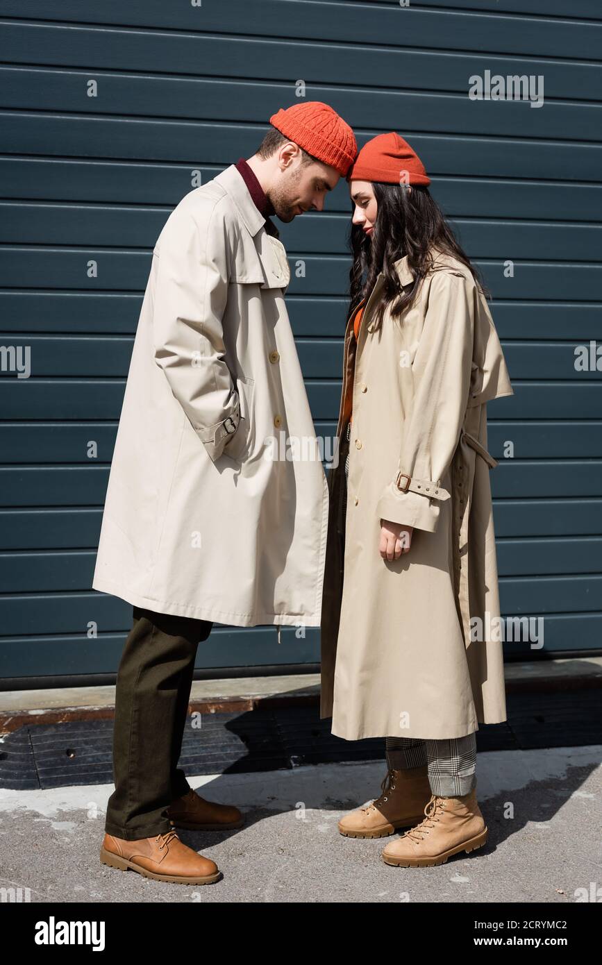 full length of stylish couple in trench coats and hats leaning on each ...