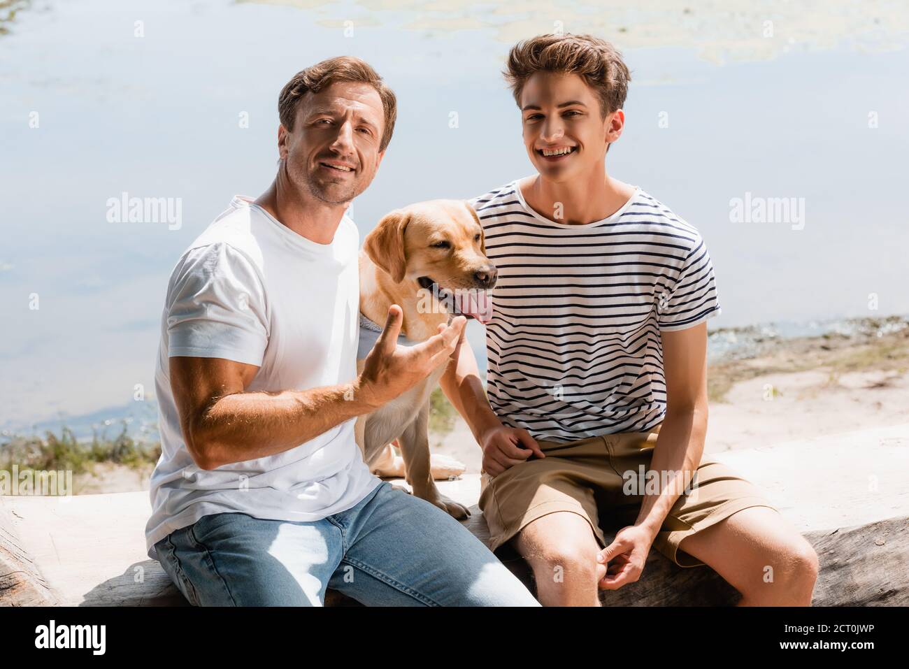 father and son sitting with golden retriever near lake Stock Photo - Alamy