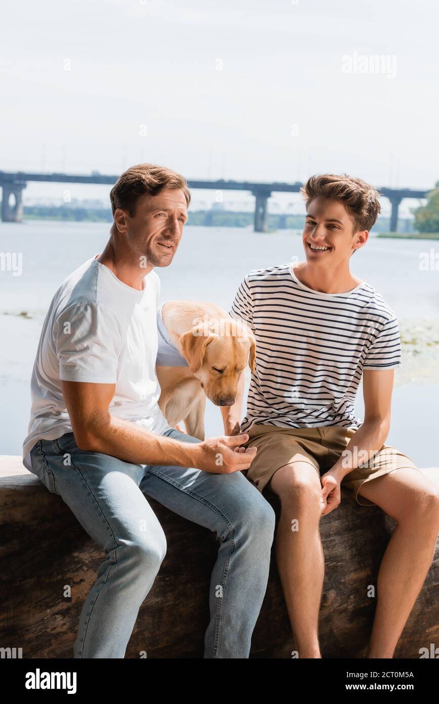 father and son sitting with golden retriever near river Stock Photo - Alamy