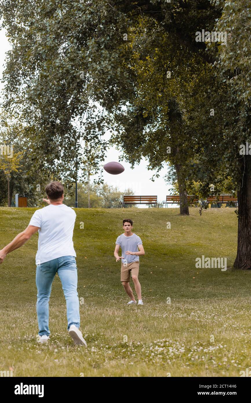 back view of father throwing rugby ball to teenager son in green park ...