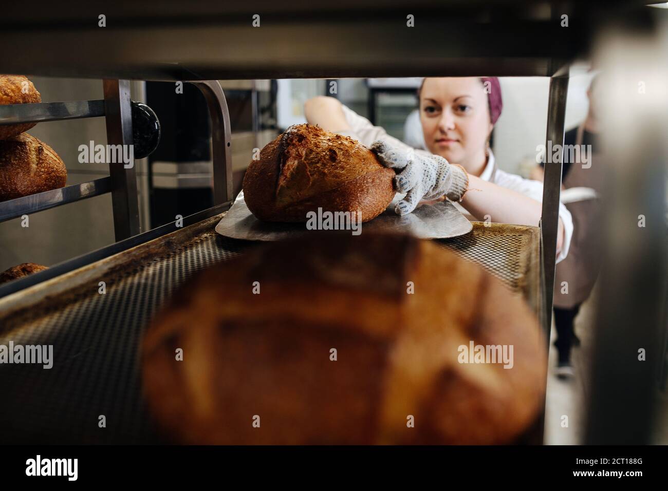 Hardworking female baker placing fresh bread on a shelf, using flat ...