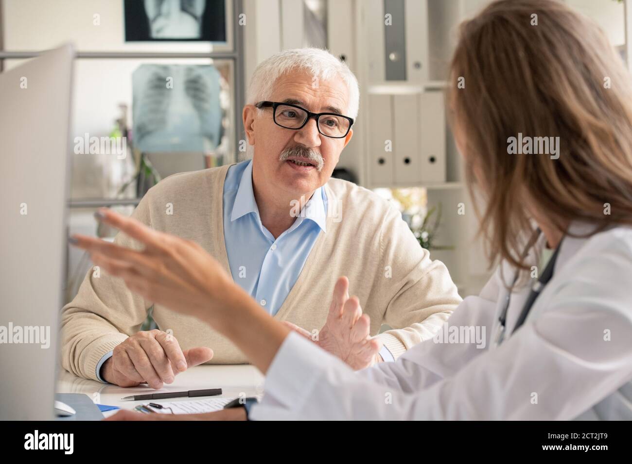 Senior patient in eyeglasses looking at doctor pointing at computer ...