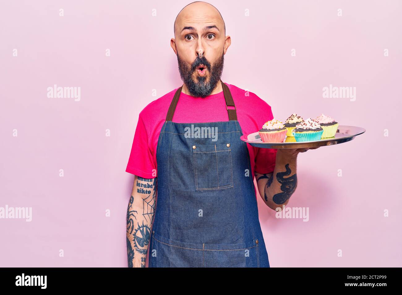 Young handsome man wearing apron holding cupcake scared and amazed with ...