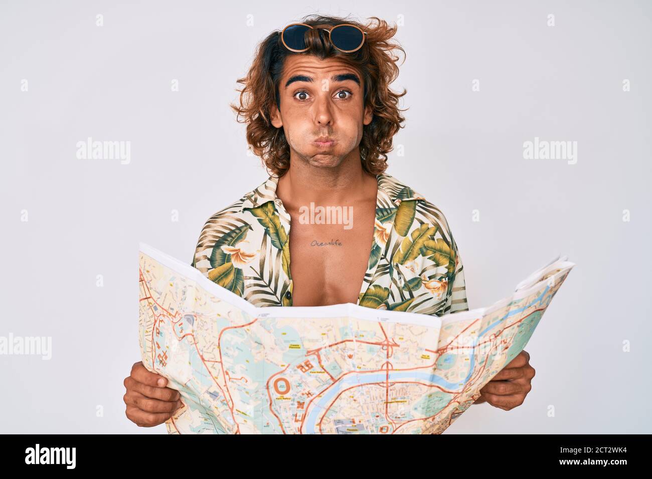 Young hispanic man wearing summer style holding map puffing cheeks with ...
