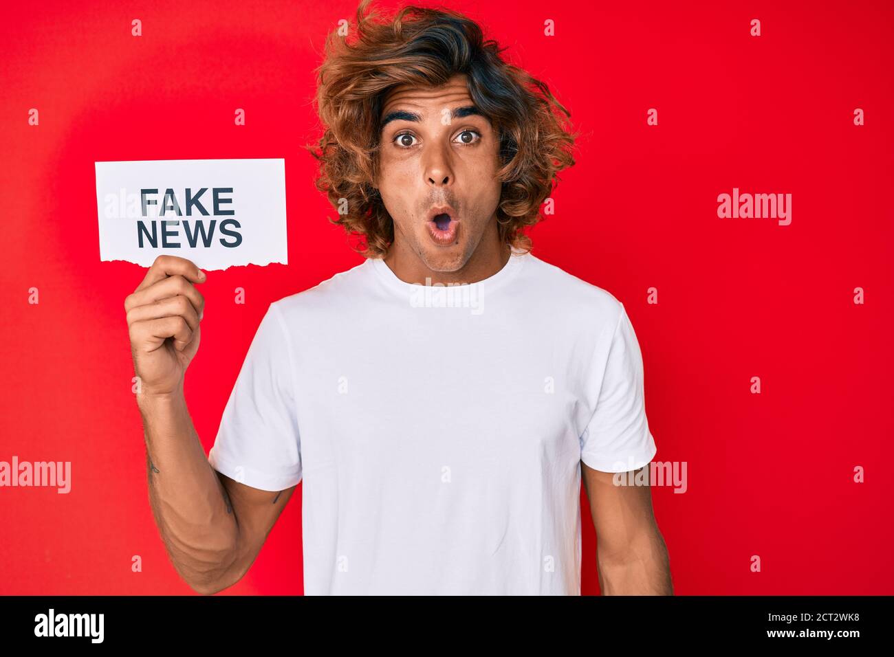 Young hispanic man holding fake news banner scared and amazed with open ...
