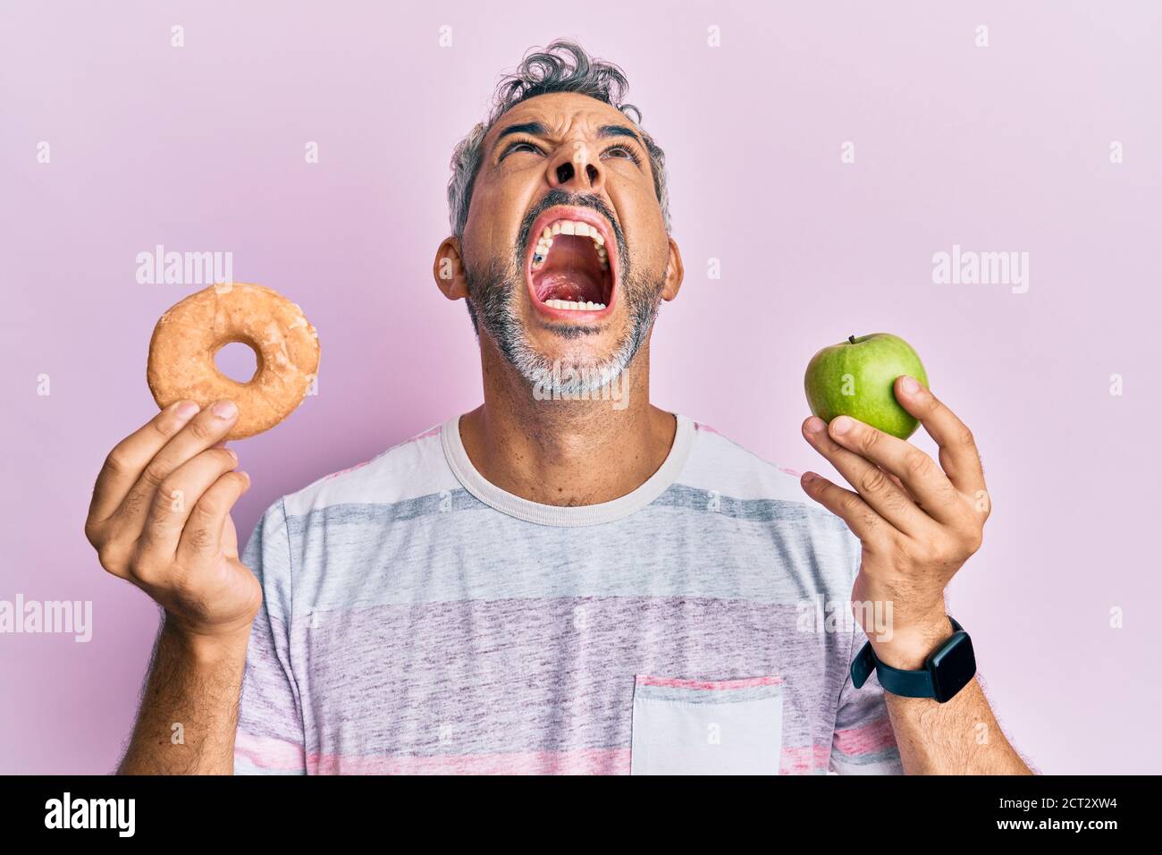 Middle age grey-haired man holding green apple and donut angry and mad ...
