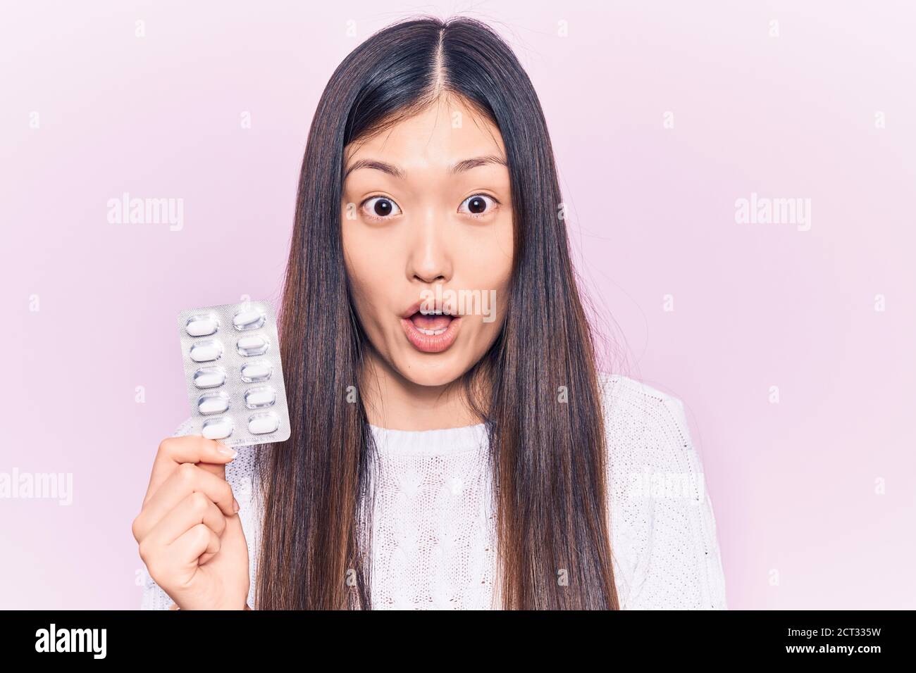 Young beautiful chinese woman holding pills scared and amazed with open ...