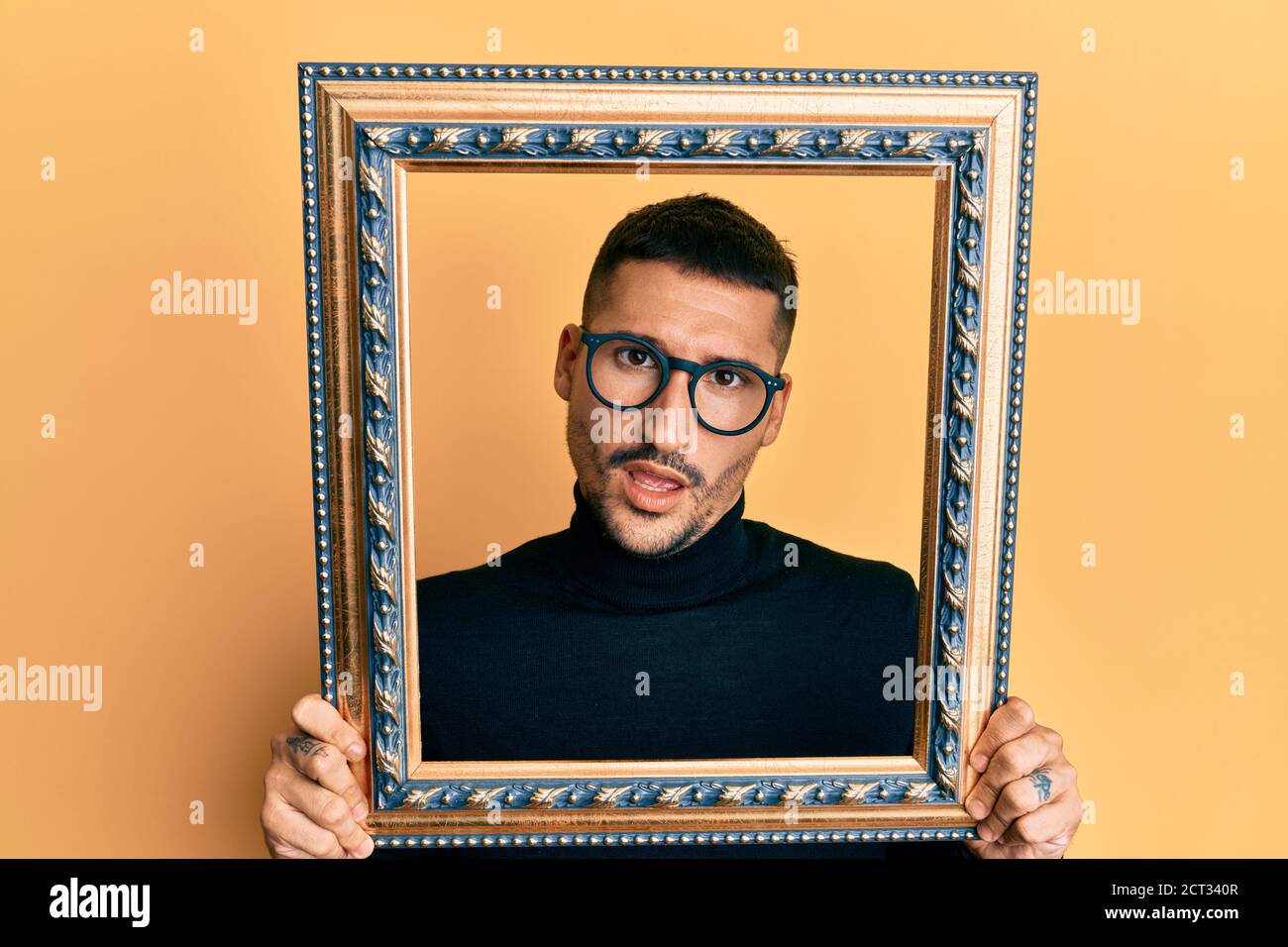 Handsome man with tattoos holding empty frame in shock face, looking ...