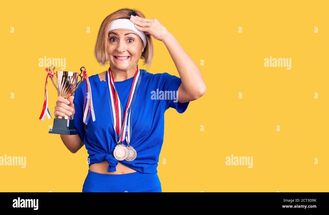 Young blonde woman holding champion trophy wearing medals stressed and ...