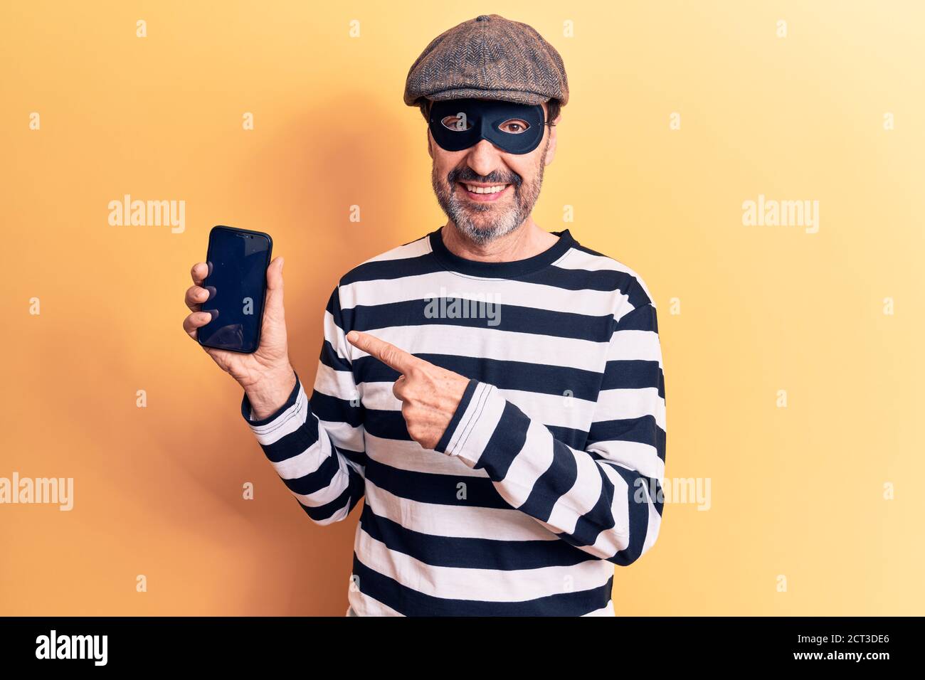 Middle age handsome burglar man wearing cap and mask holding smartphone ...