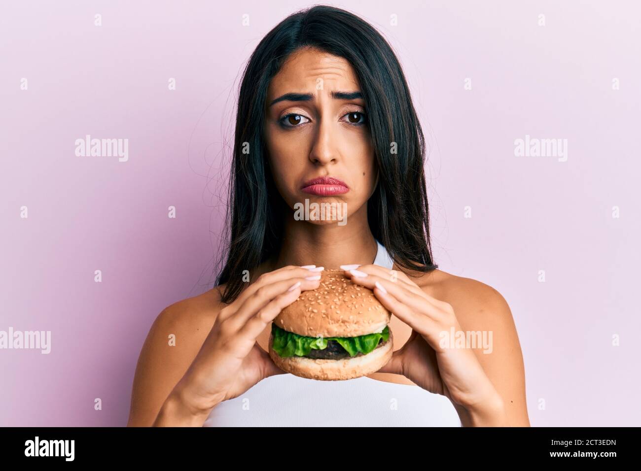 Beautiful hispanic woman eating a tasty classic burger depressed and ...