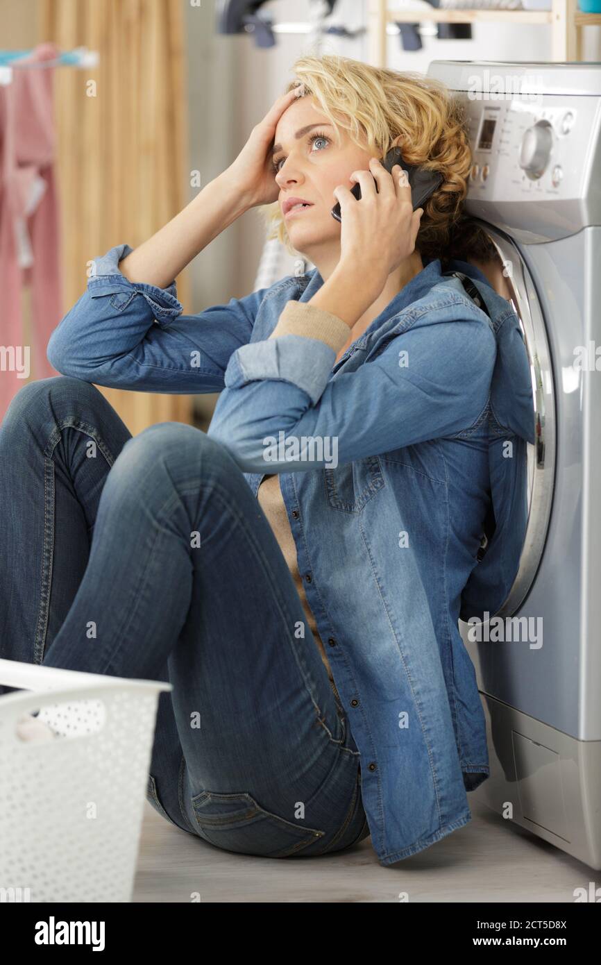 distressed woman making telephone call leaning against washing machine ...