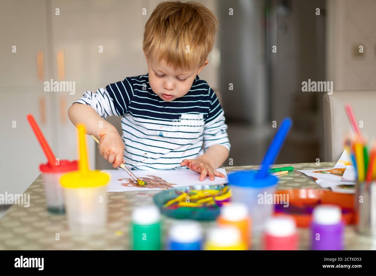 Three year old boy busy painting at home, with paint pots and brushes ...