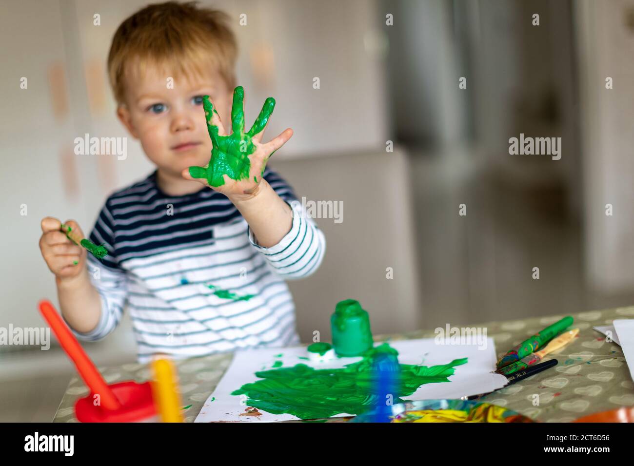 Three year old boy busy painting at home, with paint pots and brushes ...
