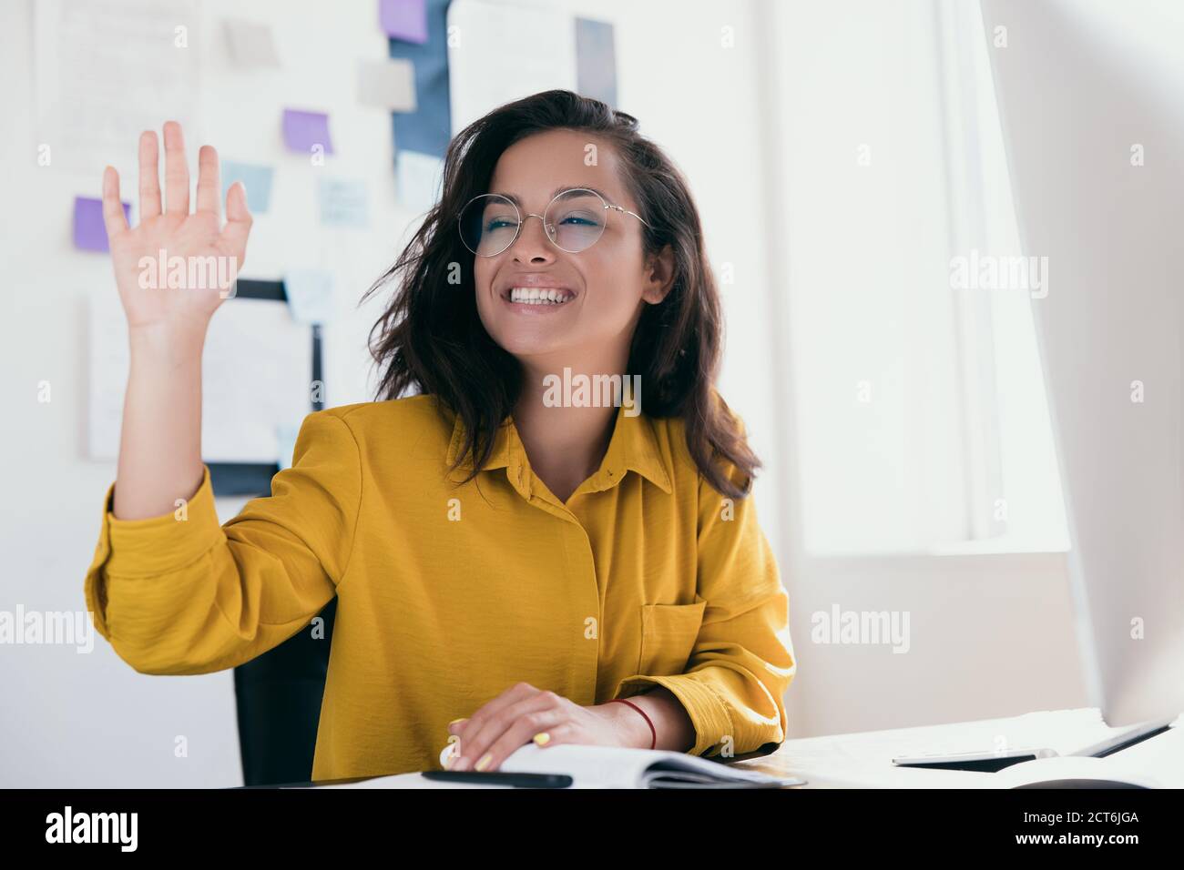 Cheerful young female office worker raised her hand up to greet someone ...