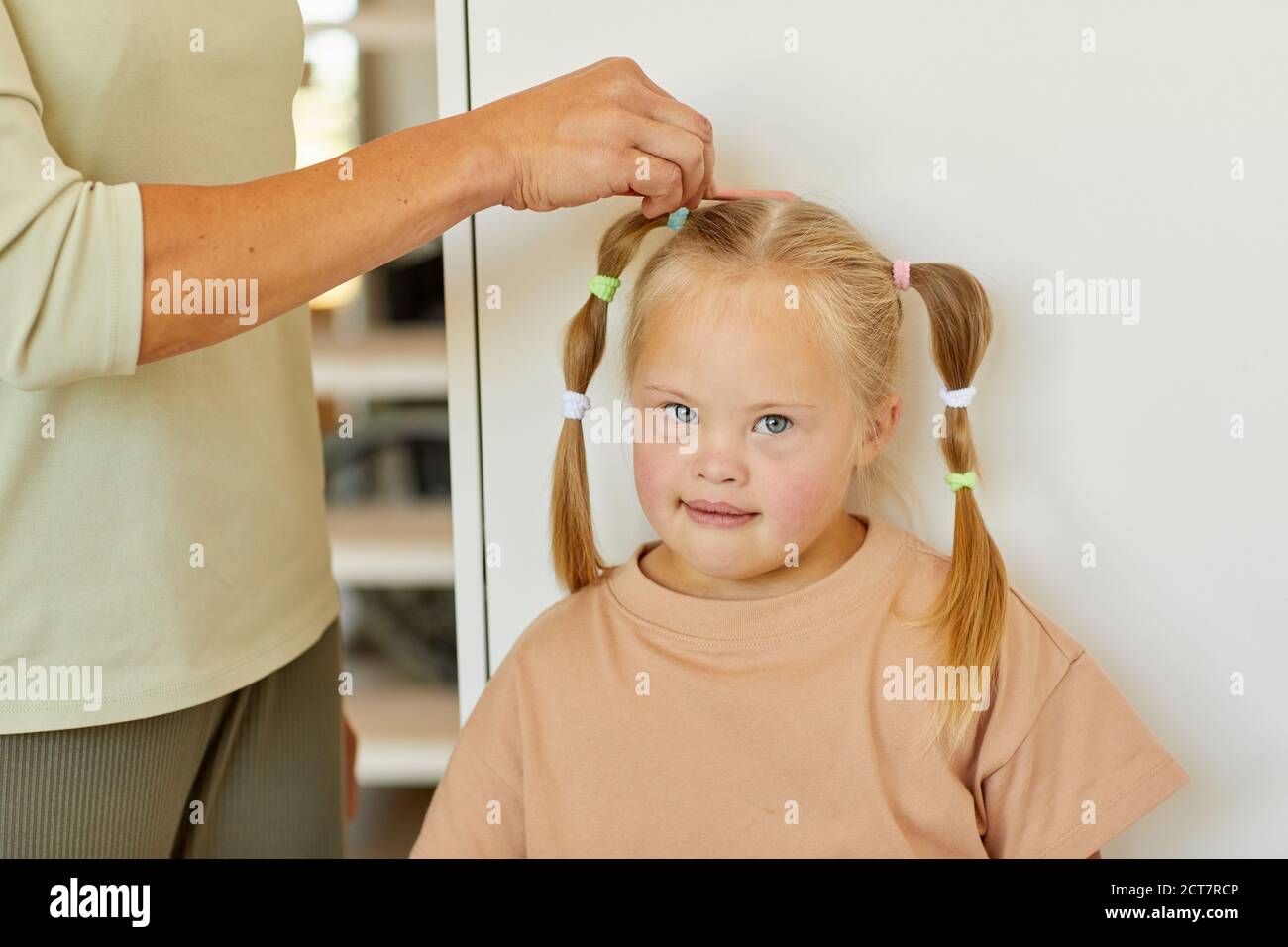 Close up of unrecognizable caring mother combing hair of cute girl with down syndrome and tying ...