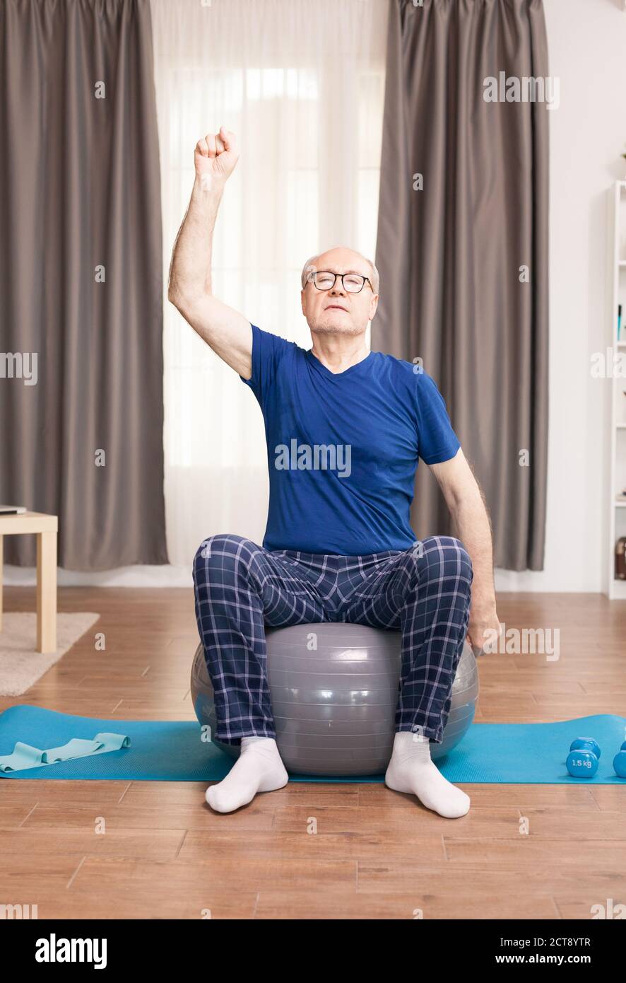 Old man training his arms sitting on stability ball in living room. Old ...