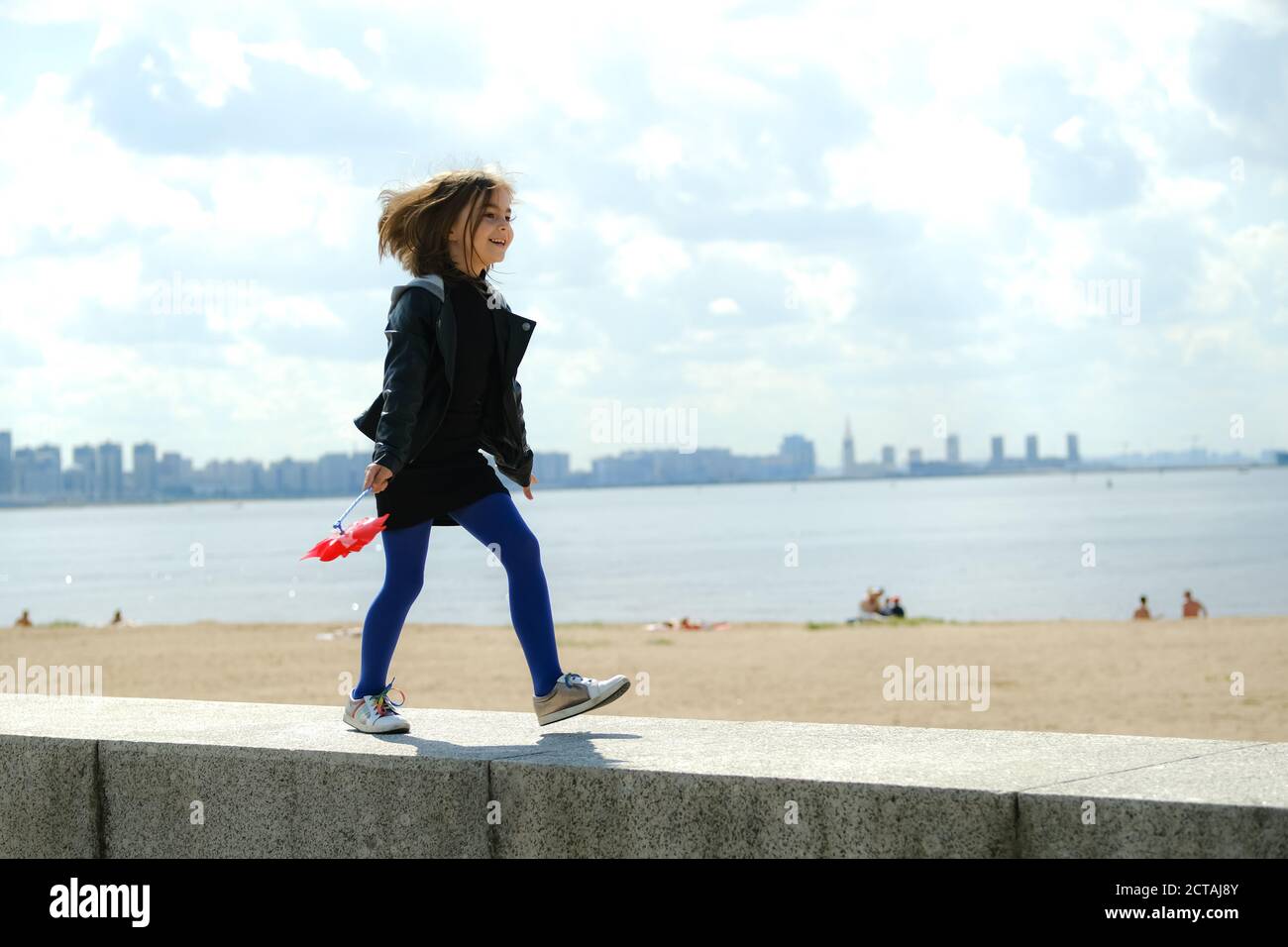 Happy girl with red wind spinner walking on the embankment on blue sky ...